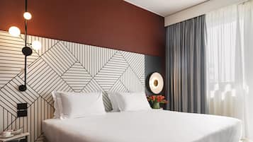 Quarto de hotel moderno com cabeceira de padrão geométrico, cama bem arrumada com lençóis brancos, mesa de cabeceira com abajur e janela com cortinas leves.