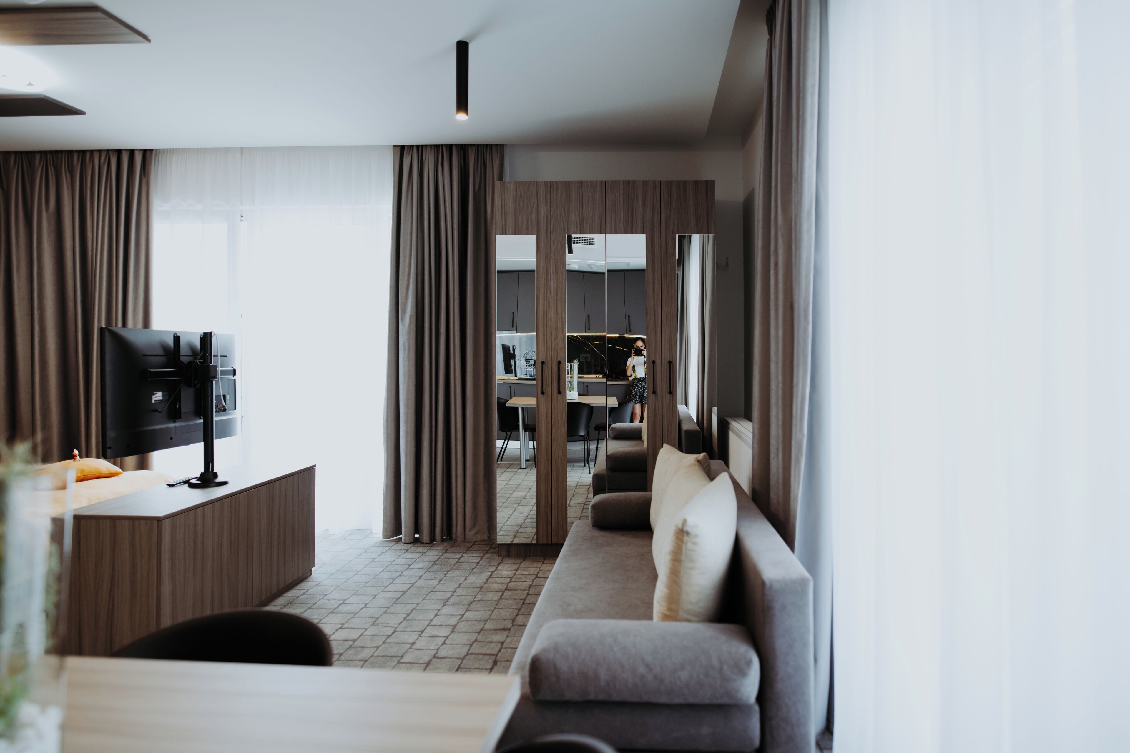 Quarto de hotel moderno com sofá, TV, escrivaninha e zona de refeições visíveis através de uma porta de vidro.