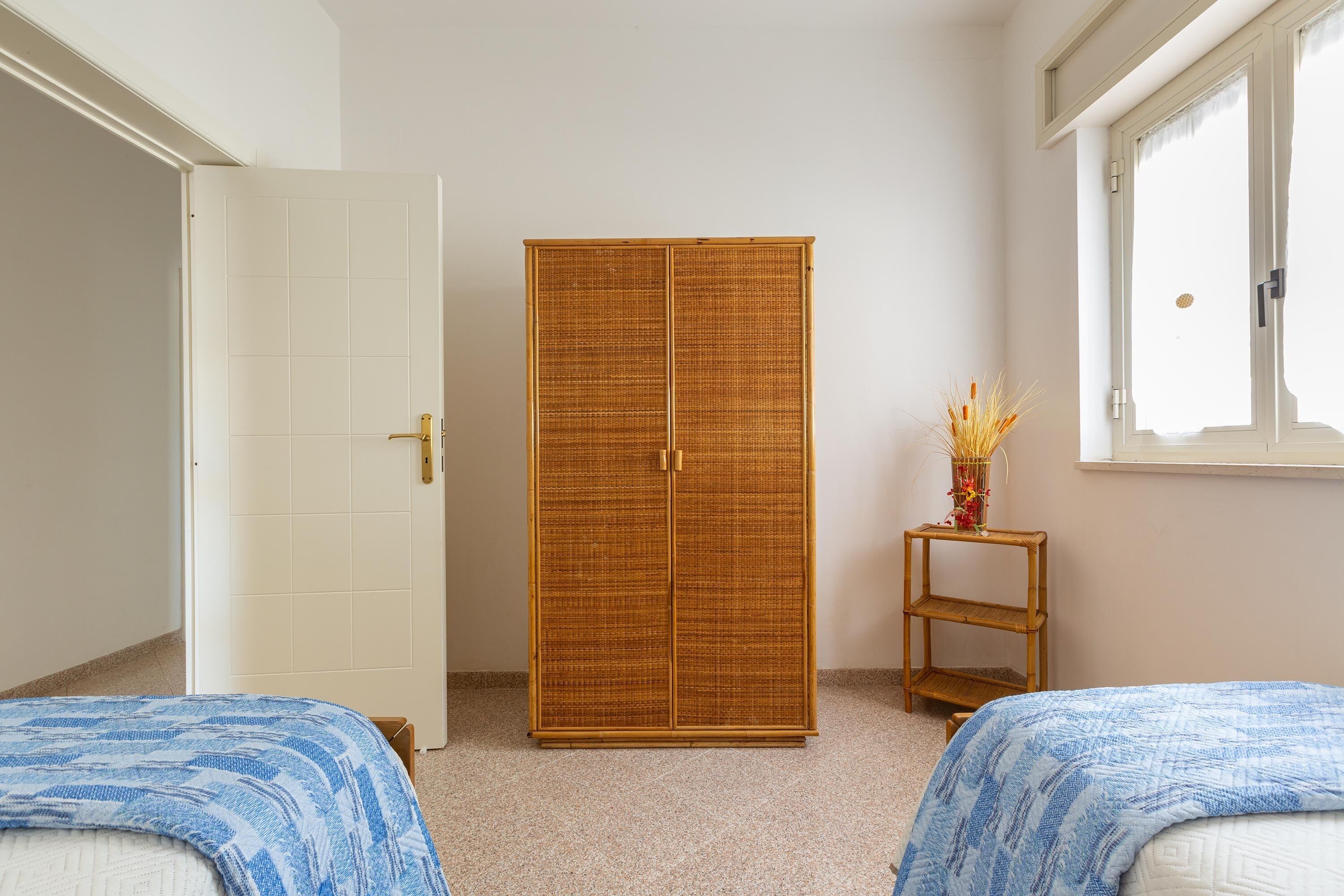 Quarto com duas camas, um guarda-roupa, uma janela e uma mesinha com um vaso.