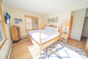 Quarto com cama de madeira, janela, mesa de cabeceira e criado-mudo.