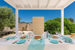 Uma área de refeições ao ar livre com uma mesa posta para dois, apresentando pizza e salada, cercada por vegetação e uma parede de pedra.