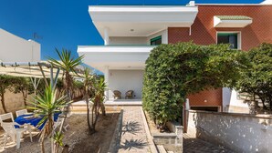 Um edifício moderno de dois andares com área de estar coberta ao ar livre, um caminho pavimentado e um pequeno jardim com plantas.