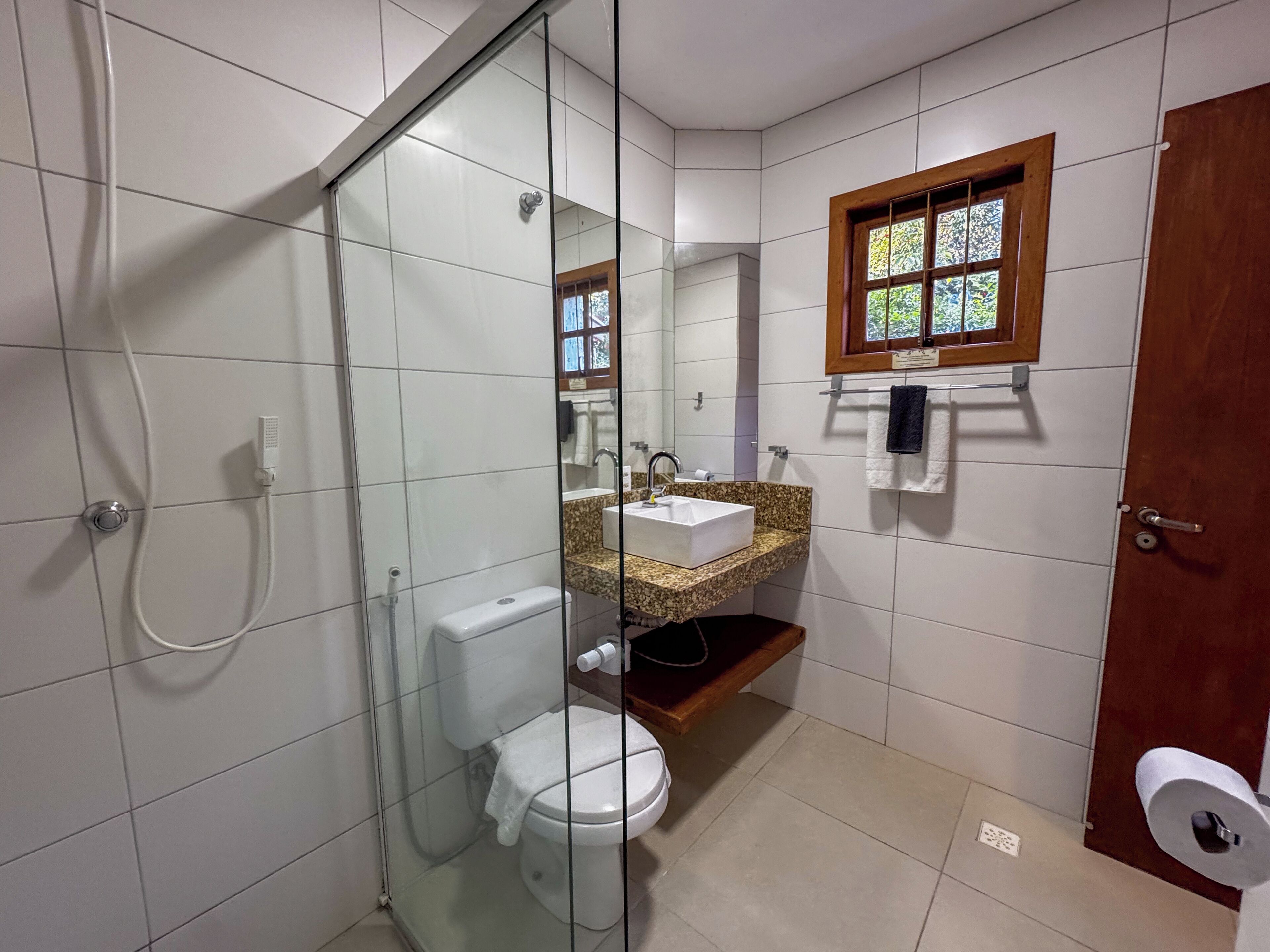 Uma casa de banho com cabine de duche em vidro, sanita, lavatório com bancada de madeira e janela com estrutura de madeira.