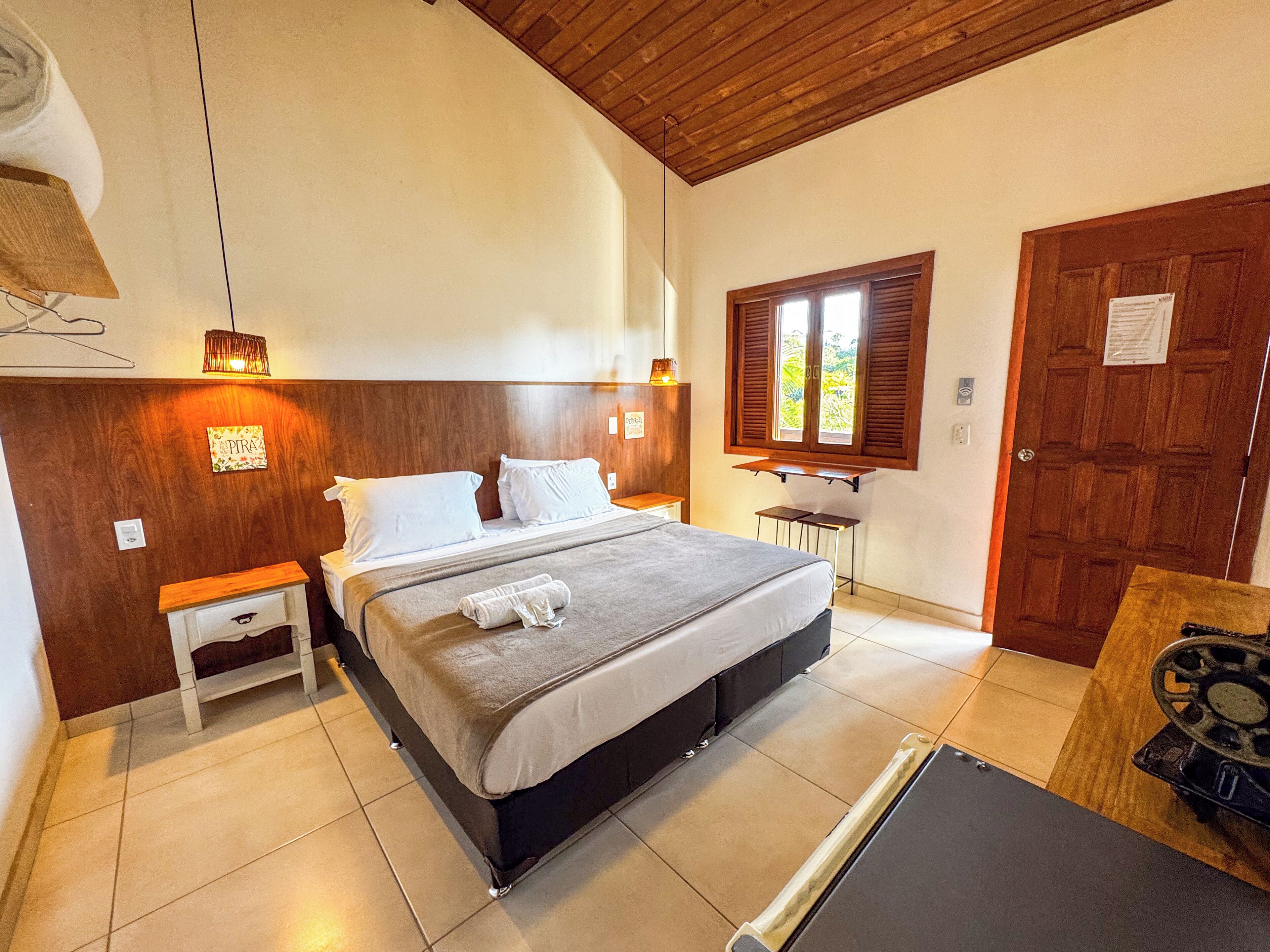 Um quarto com cama, mesas de cabeceira, teto de madeira, janela com persianas e uma porta.