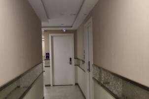 Corredor com porta no final, corrimão à esquerda e luminária fixada na parede.