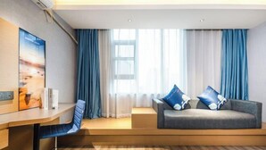 Quarto de hotel moderno com escrivaninha de madeira, sofá cinza com almofadas azuis e uma janela grande com cortinas azuis.