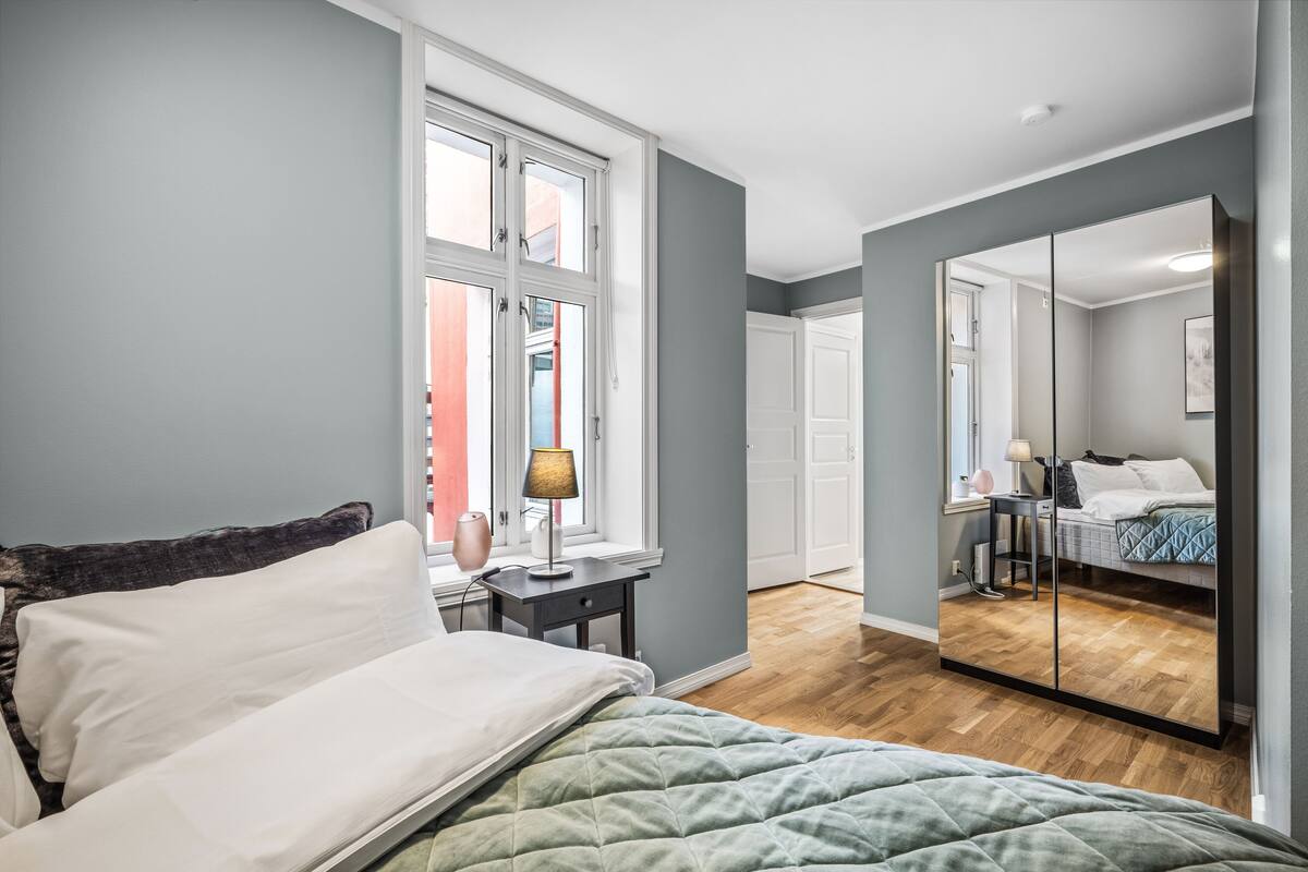 Quarto com cama, mesa de cabeceira com abajur, espelho e vista para outro cômodo através de uma porta.