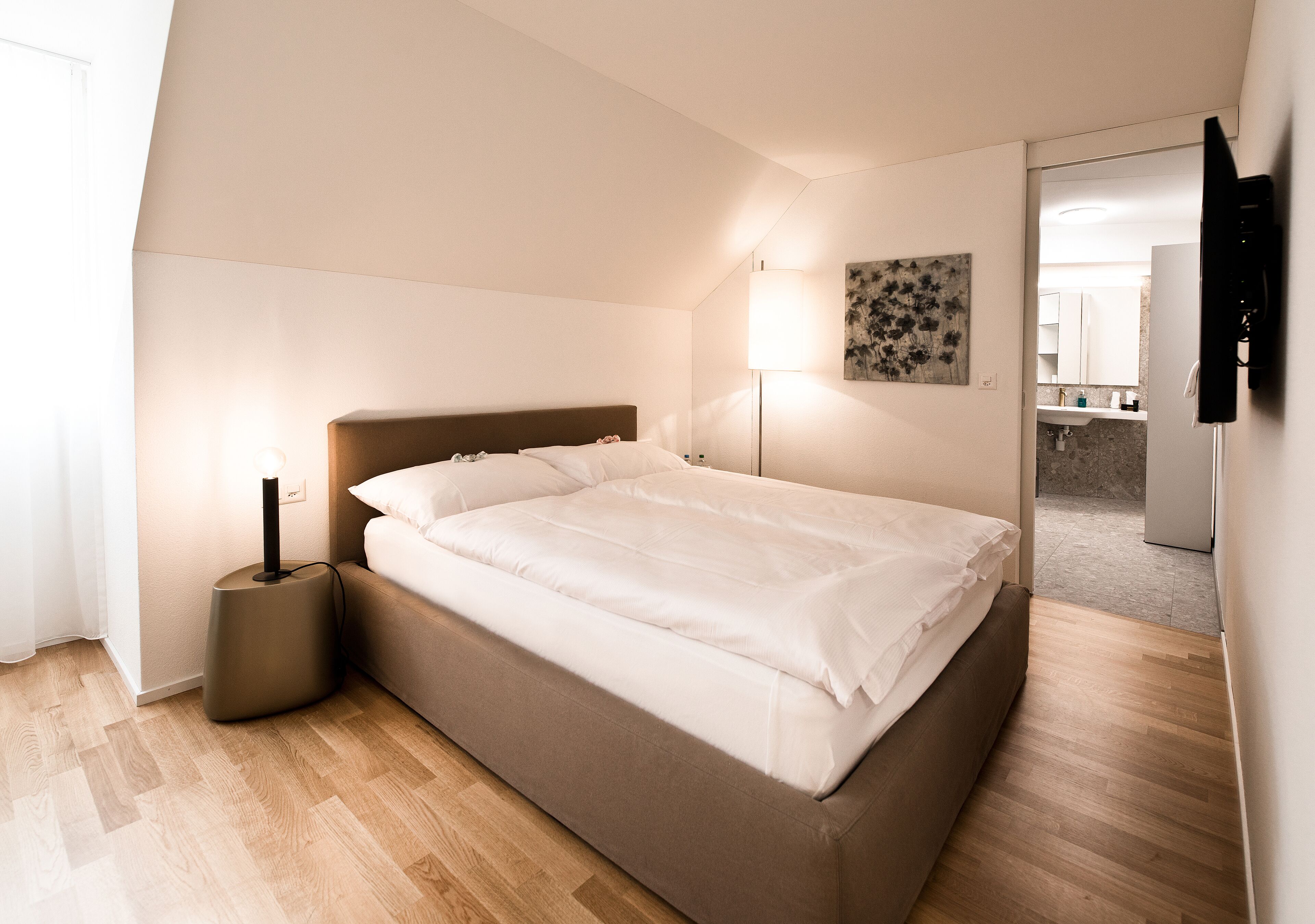 Quarto bem arrumado com cama, abajur e televisão presa à parede.