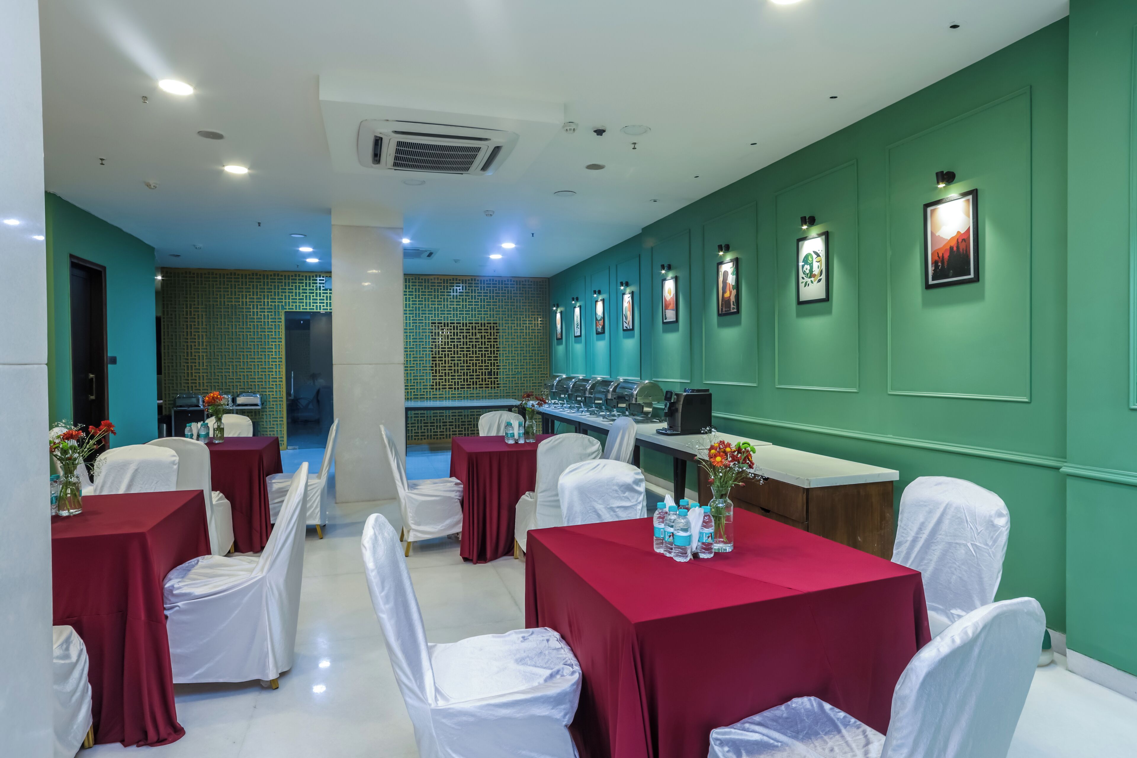 Uma sala de jantar com mesas cobertas por toalhas vermelhas e cadeiras brancas, uma parede verde decorativa e quadros nas paredes.
