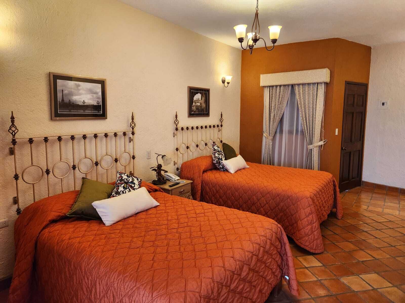 Quarto de hotel com duas camas, cada uma com colcha acolchoada vermelha e almofadas a combinar. Há duas quadros na parede, um candelabro e uma porta com cortinas.