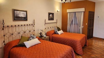 Quarto de hotel com duas camas, cada uma com colcha acolchoada vermelha e almofadas a combinar. Há duas quadros na parede, um candelabro e uma porta com cortinas.