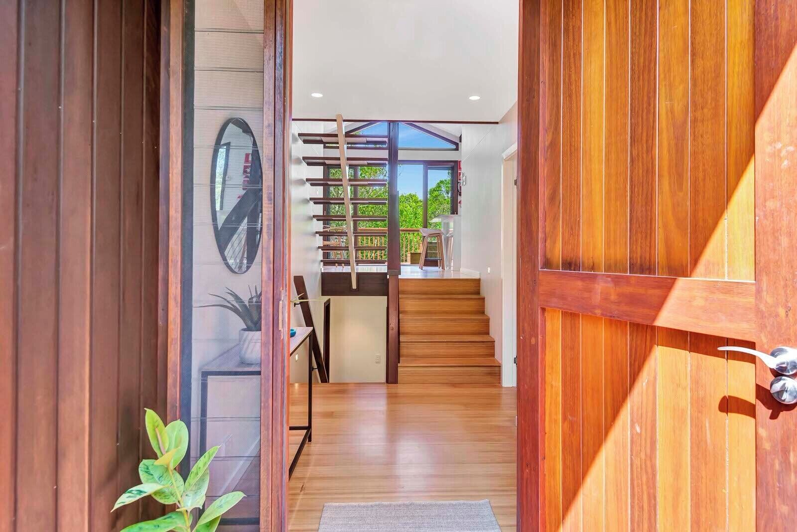 Um interior moderno com uma escada de madeira que leva a uma varanda, uma porta de madeira grande e vista para o verde lá fora.