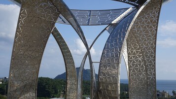 Estrutura metálica com padrão geométrico, possivelmente parte de uma ponte ou elemento arquitetônico grandioso, com vista para uma paisagem natural com árvores e um corpo d'água.
