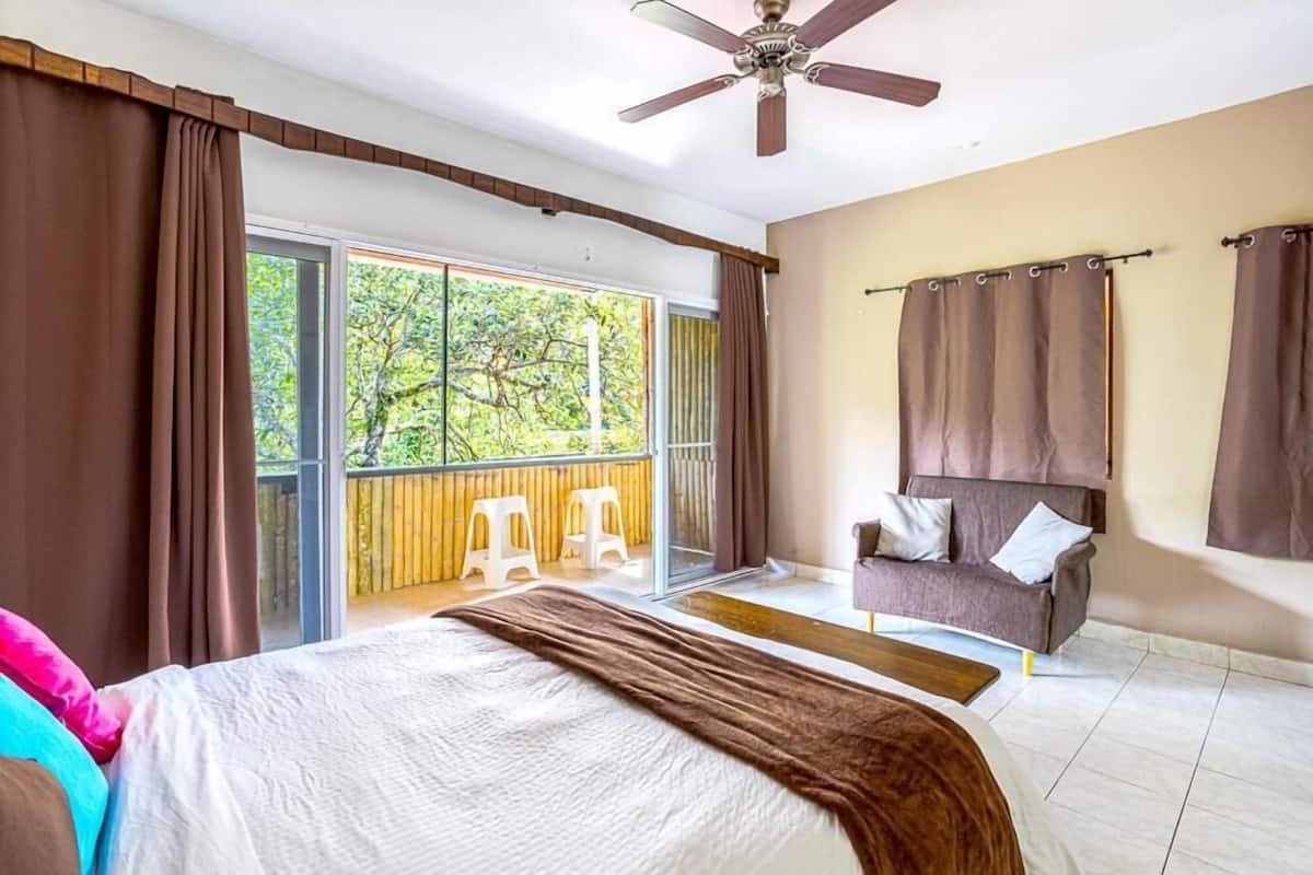 Quarto com cama grande, sofá, ventilador de teto e varanda com vista para o verde.