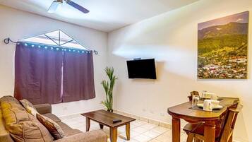 Sala de estar com sofá, mesa de centro, mesa de jantar com cadeiras, ventilador de teto, TV fixada na parede e janela com cortinas.