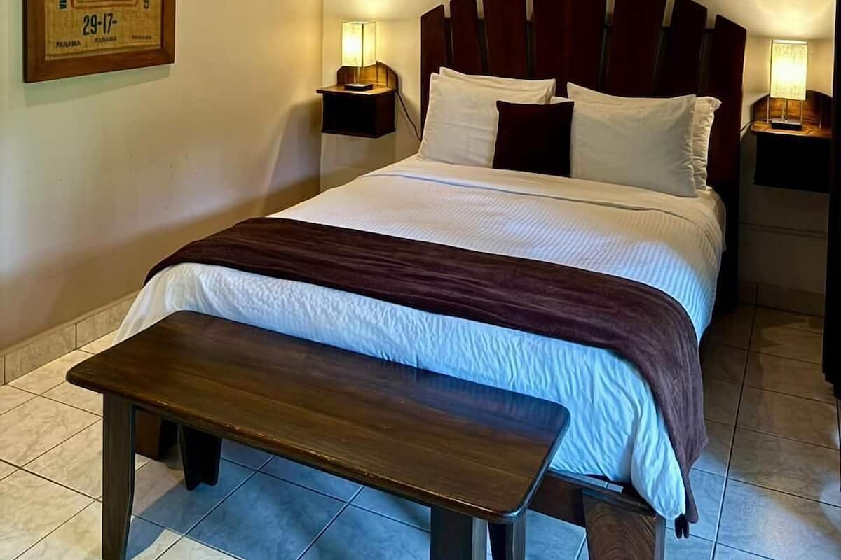 Quarto de hotel com cama, mesas de cabeceira com luminárias, ventoinha no teto, quadro na parede e um banco de madeira.