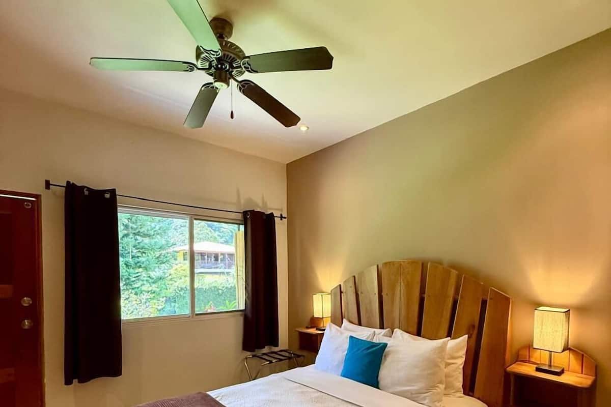 Quarto de hotel com uma cama, ventoinha no teto, janela com cortinas, mesa de cabeceira com abajur e cabeceira de madeira.