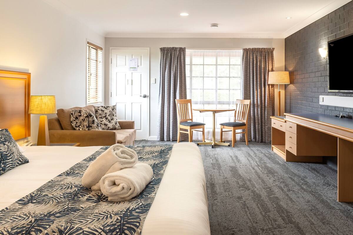 Quarto de hotel com uma cama grande, uma escrivaninha, uma cadeira, uma televisão e uma janela com cortinas.