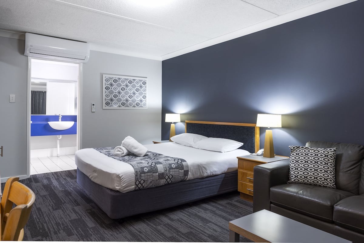 Quarto de hotel com cama, escrivaninha, cadeira, sofá, mesa de cabeceira, luminária, uma obra de arte na parede e banheiro visível por uma porta aberta.