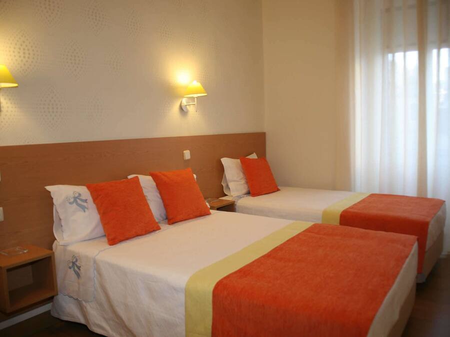 Quarto de hotel com duas camas, cada uma com almofadas laranja e colcha branca. Há duas luminárias fixadas na parede, uma de cada lado das camas.
