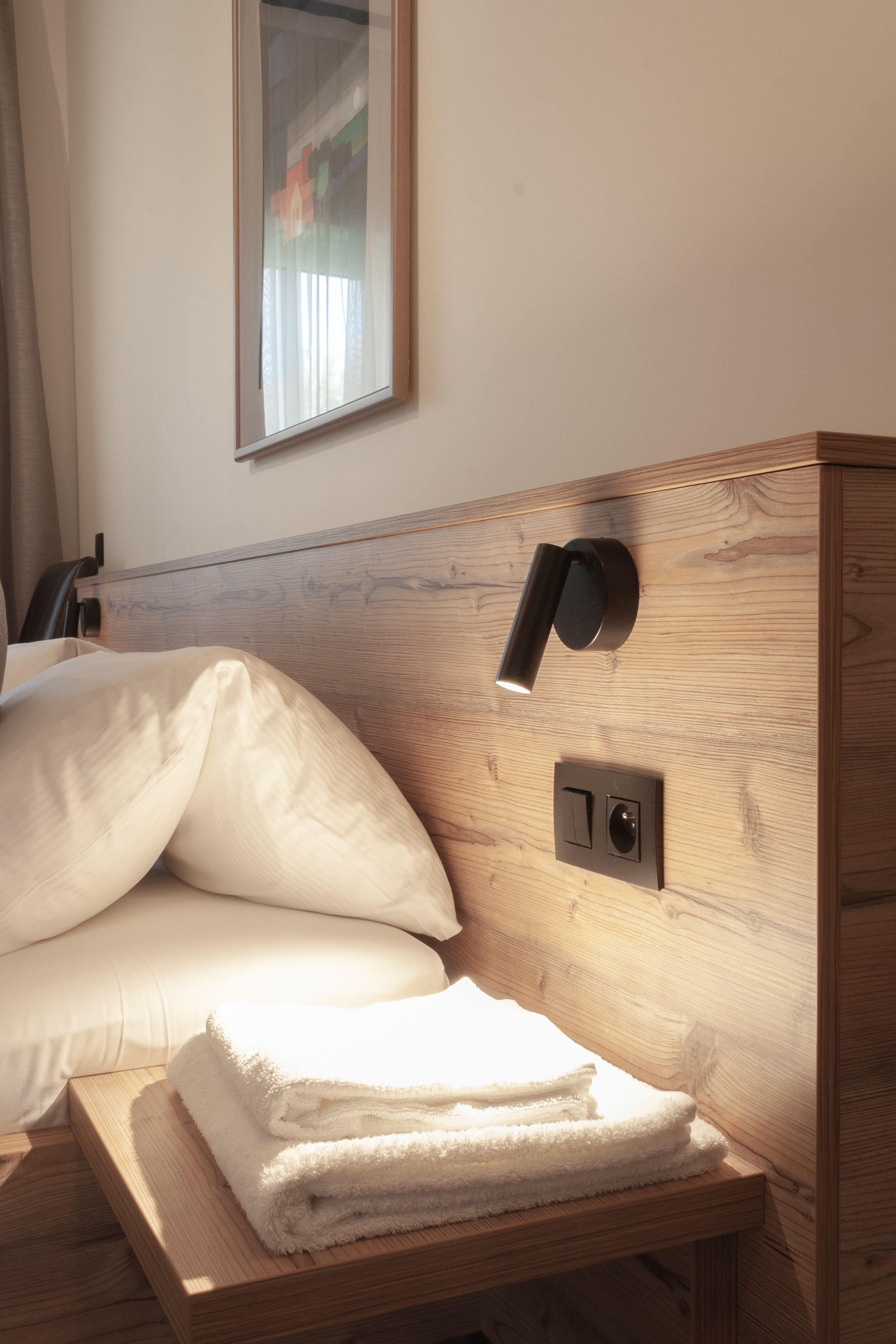 Uma cama bem arrumada com lençóis brancos e uma toalha dobrada sobre uma mesa de cabeceira de madeira.