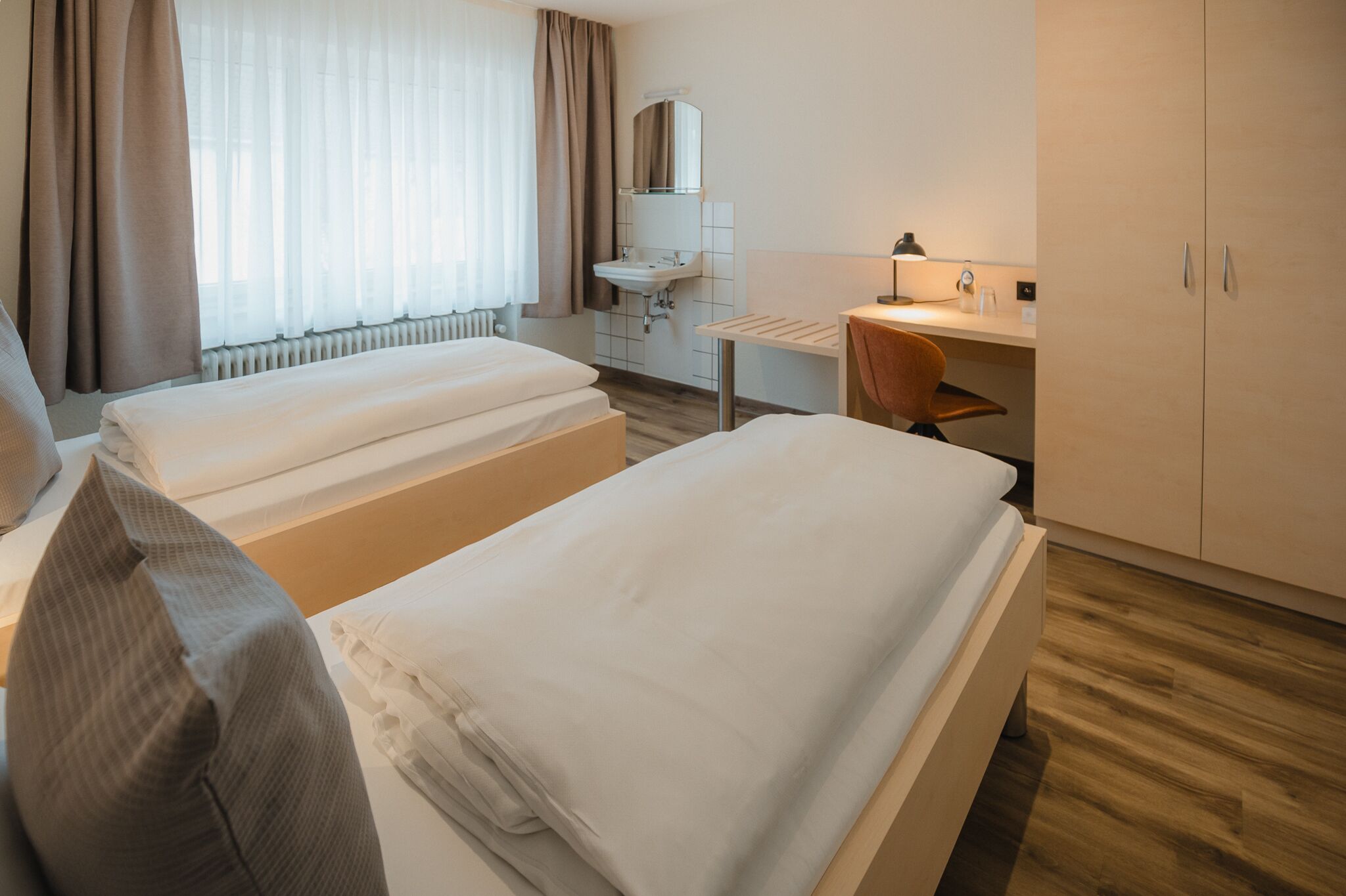 Quarto de hotel com duas camas, uma escrivaninha, uma cadeira, banheiro com pia e espelho, e um guarda-roupa.