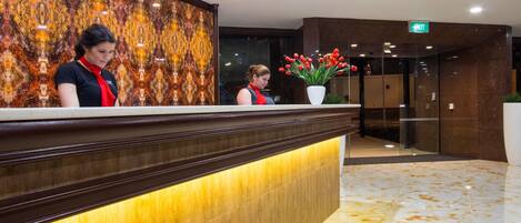 Uma área de recepção de hotel com piso de mármore, balcão de atendimento e uma planta em vaso com flores vermelhas.
