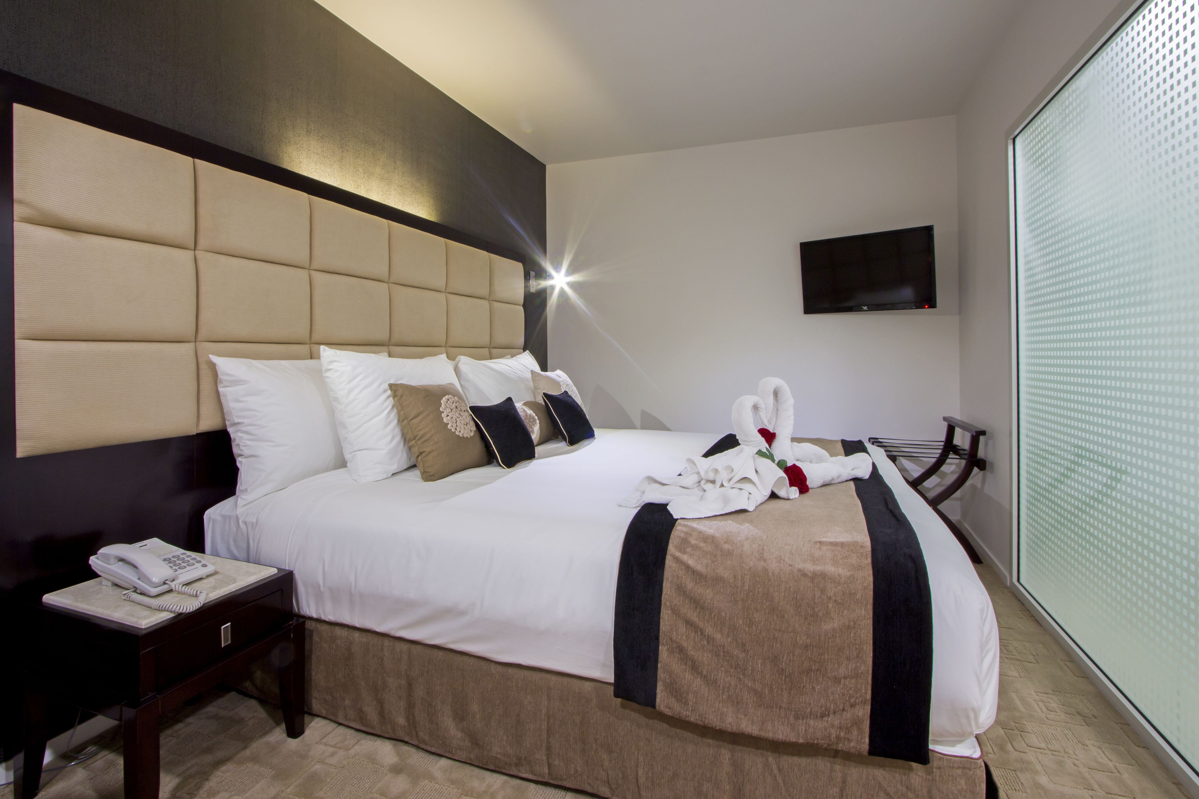 Quarto de hotel com uma cama grande, mesa de cabeceira com telefone, televisão de tela plana na parede e uma janela ampla com persianas.