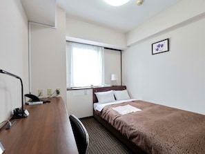 Quarto de hotel com uma cama de solteiro, uma mesa com telefone, uma luminária e uma janela com cortinas.