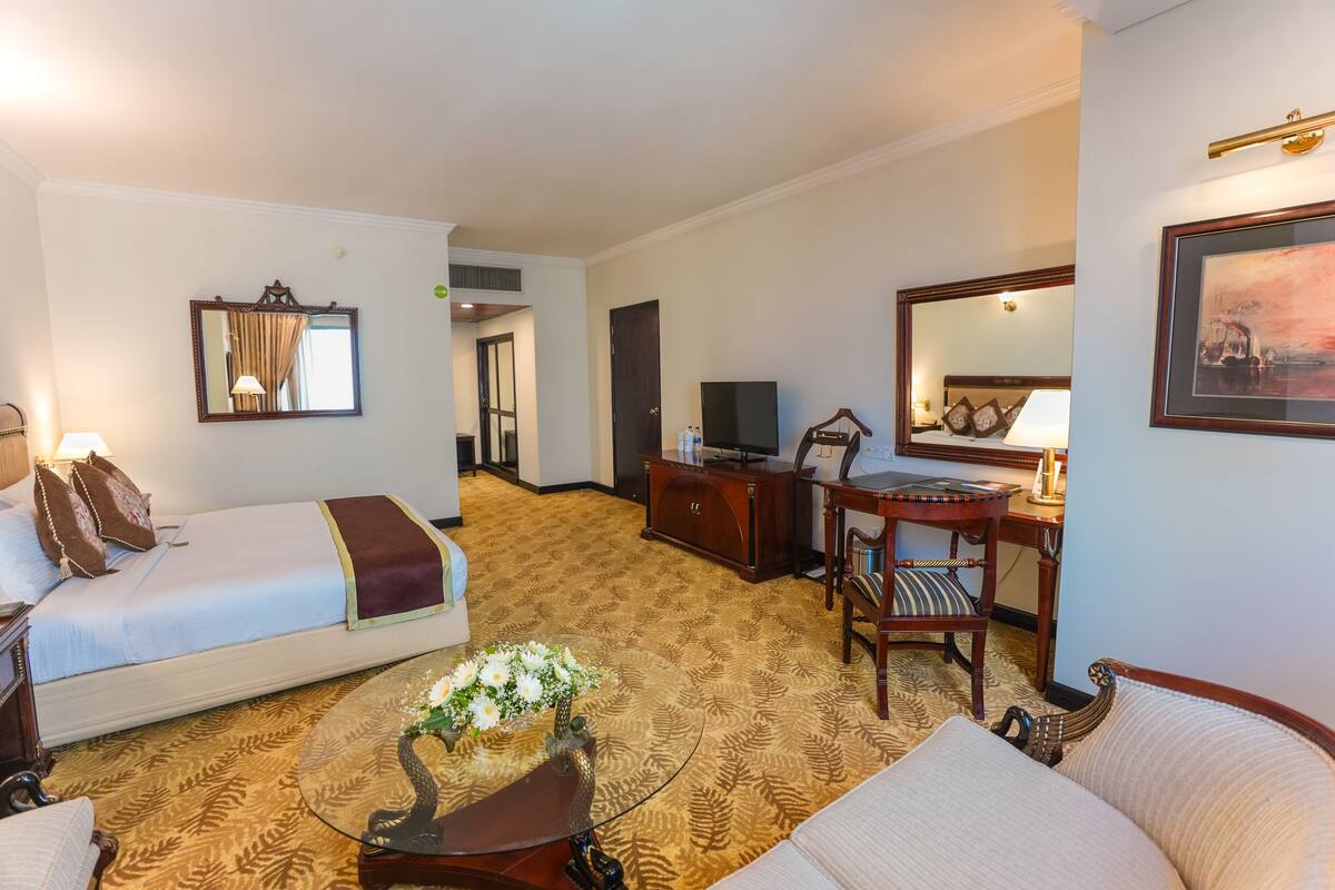 Quarto de hotel com cama, sofá, mesinha com flores, televisão, escrivaninha e cadeira.