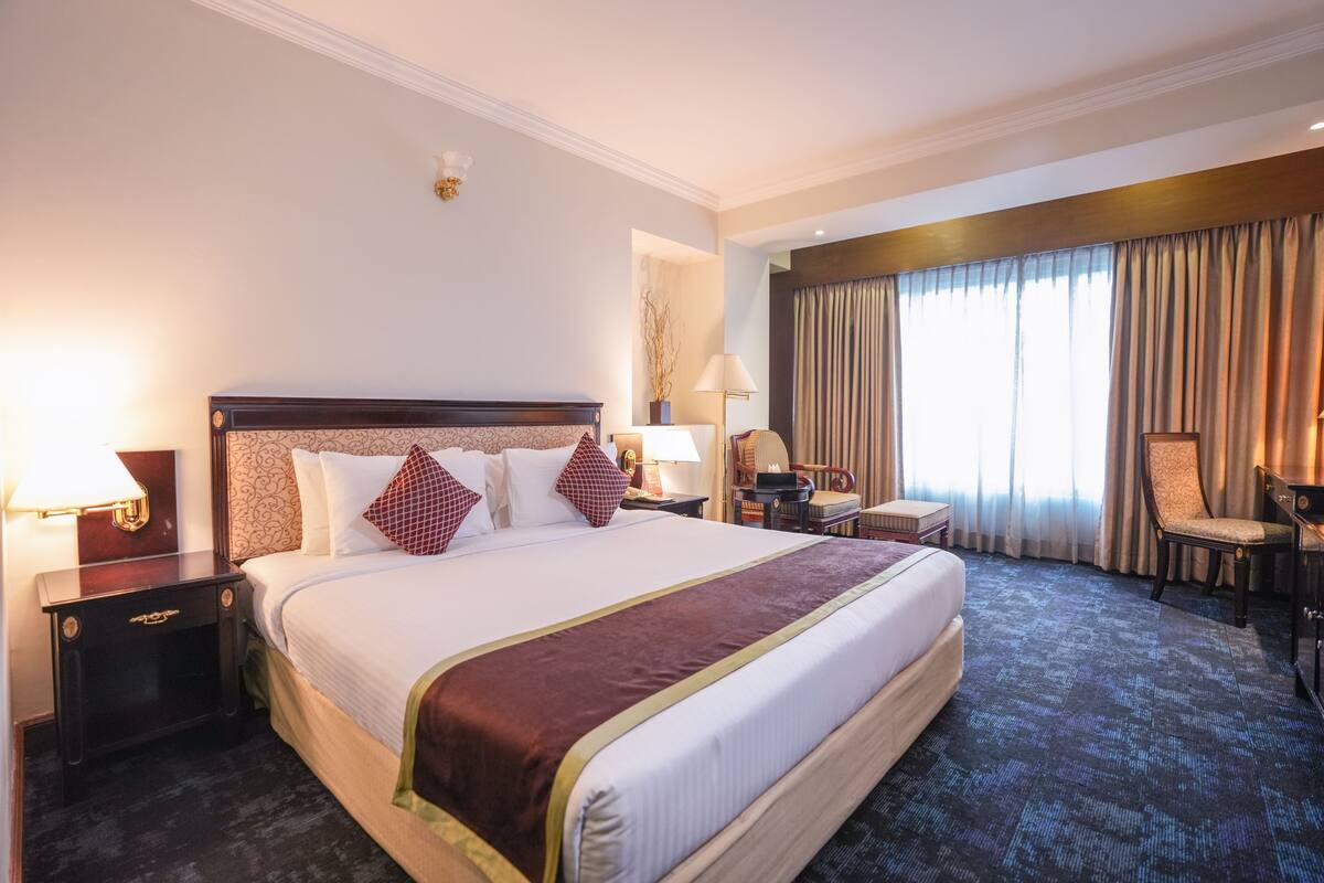 Quarto de hotel com uma cama grande, mesa de cabeceira, escrivaninha, cadeira e janela com cortinas.