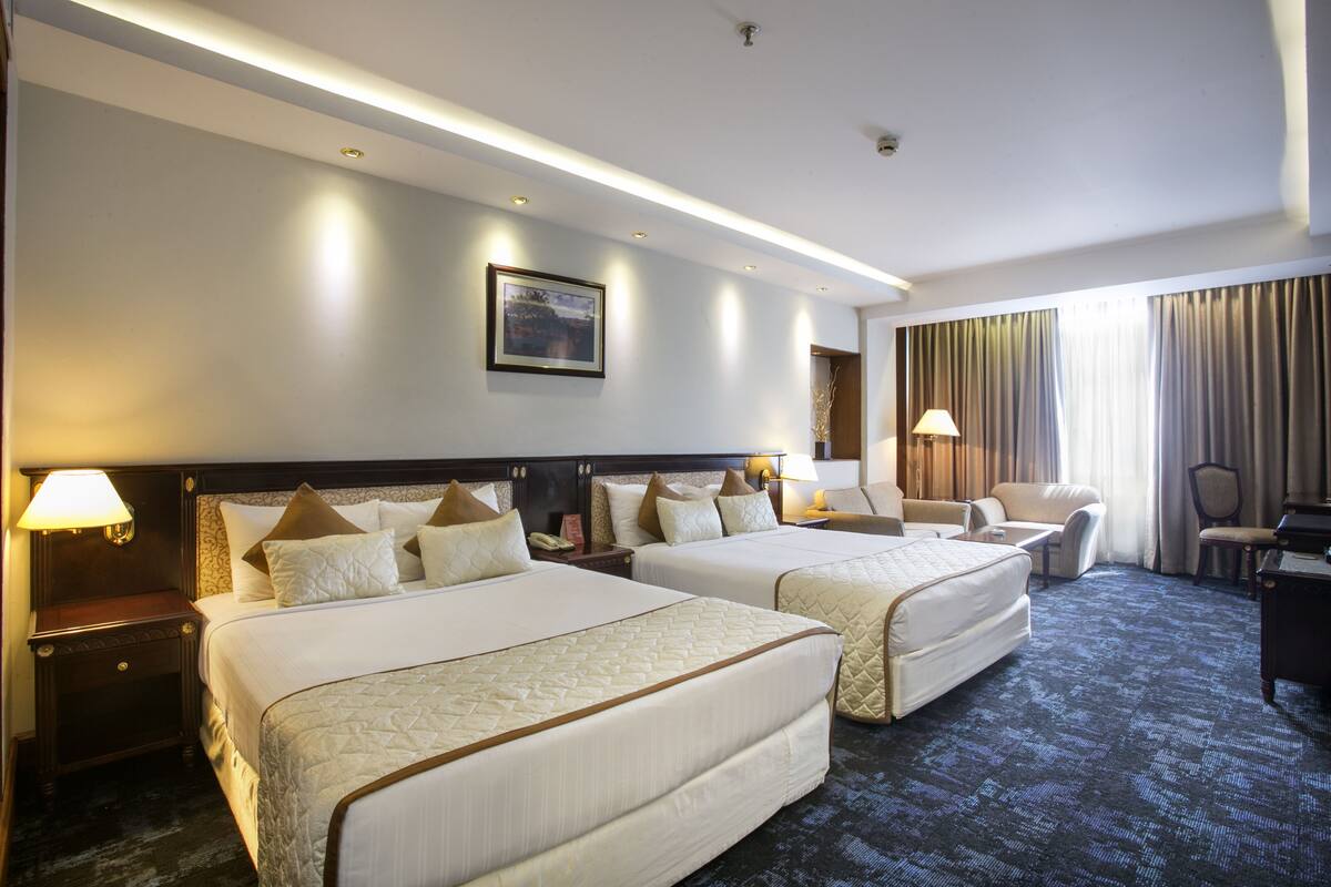 Quarto de hotel com duas camas, uma escrivaninha, uma cadeira e uma janela grande com cortinas.