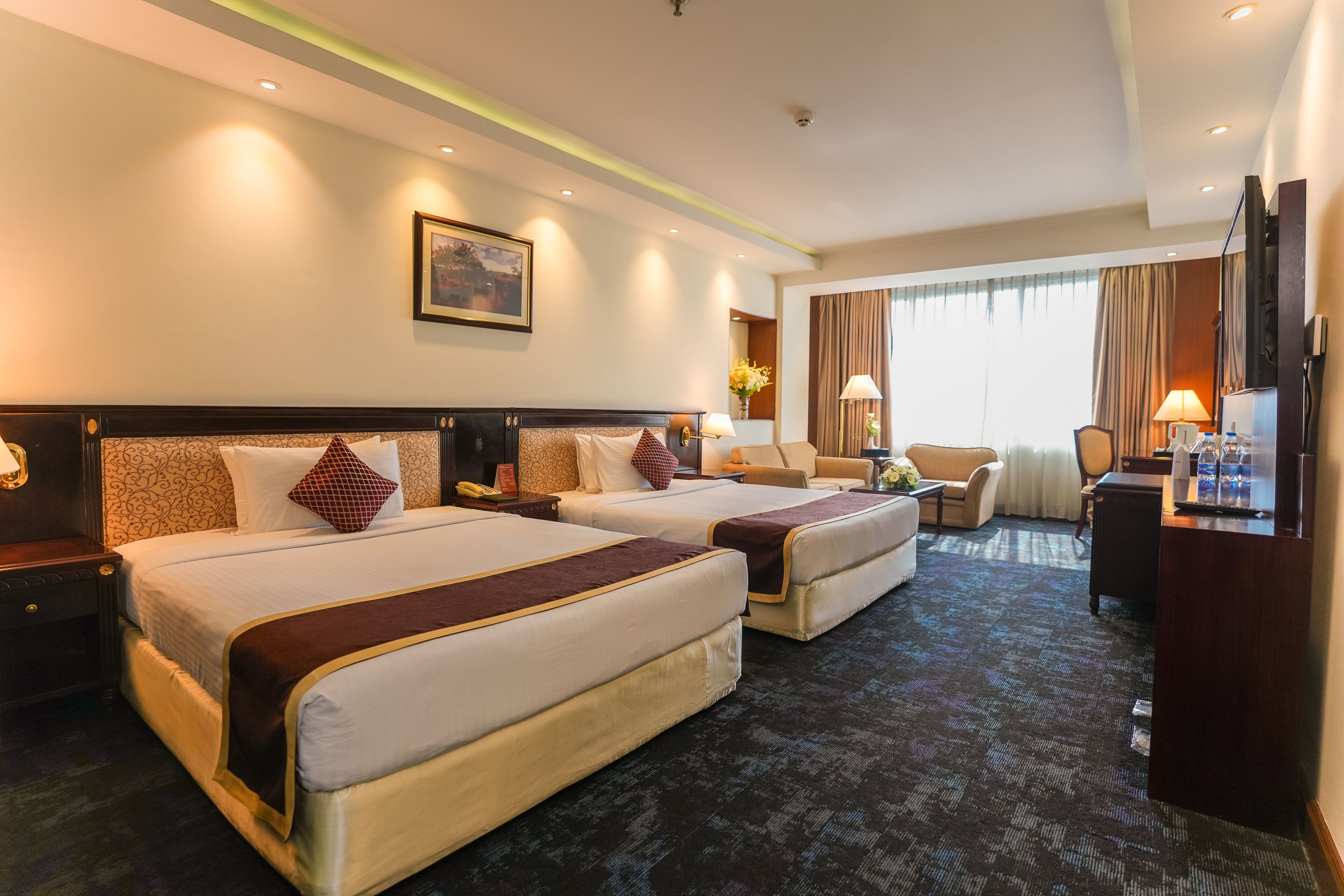 Quarto de hotel com duas camas, uma escrivaninha, uma cadeira e uma janela grande com cortinas.
