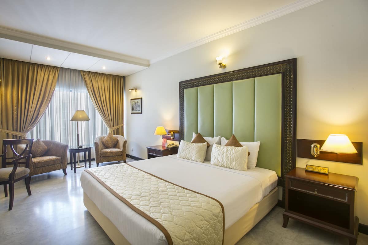 Quarto de hotel com uma cama grande, duas poltronas, uma mesa de cabeceira com abajur e uma janela com cortinas.