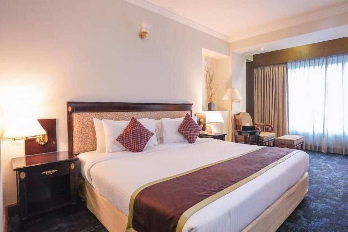 Quarto de hotel com cama grande, mesas de cabeceira, escrivaninha, cadeira e vista para o exterior.