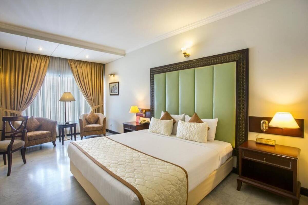 Quarto de hotel com uma cama grande, duas cadeiras, uma mesa de cabeceira, um abajur e uma janela com cortinas.