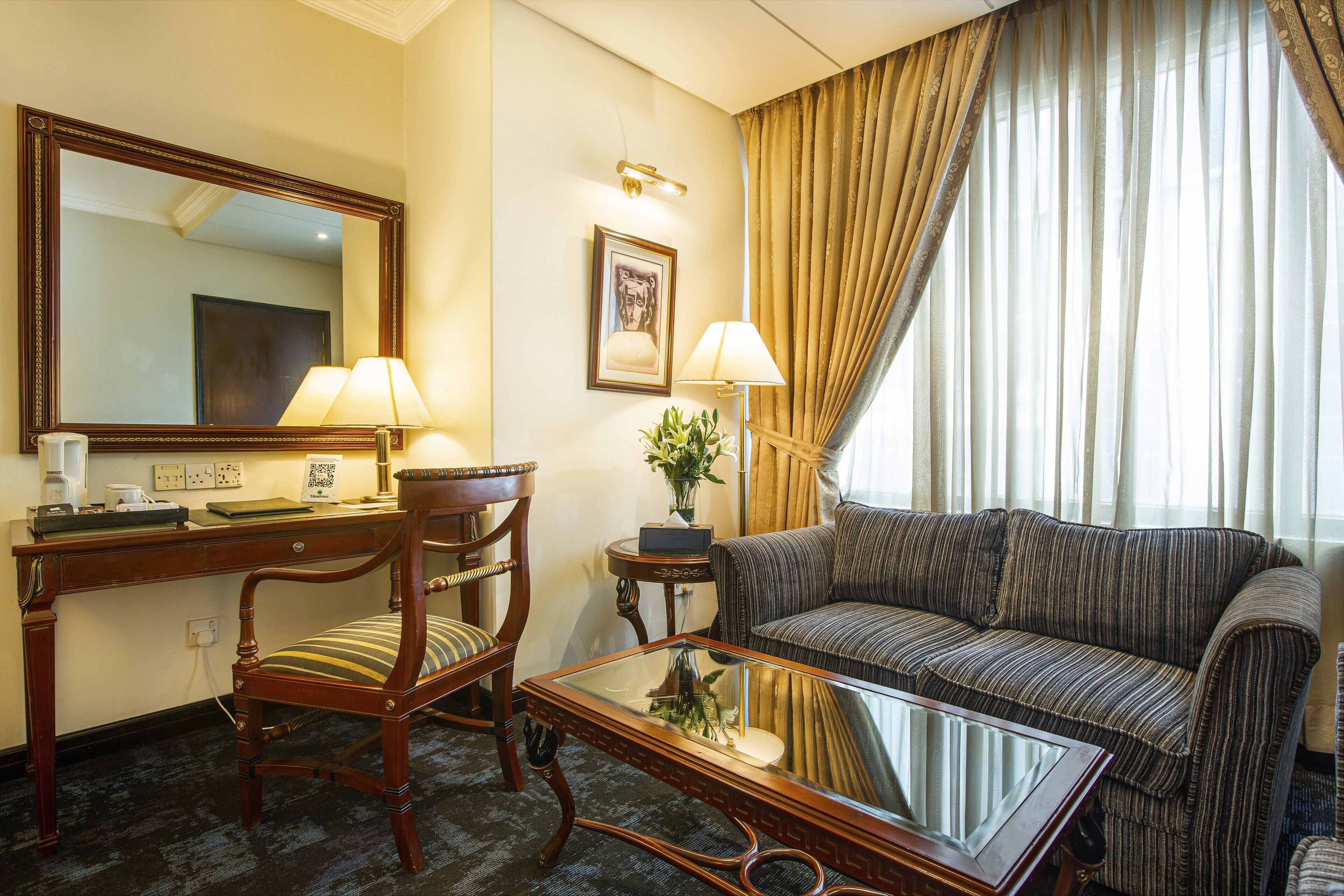Quarto de hotel com escrivaninha de madeira, sofá riscado, mesa de centro de vidro, uma cadeira, espelho e um quadro na parede.