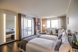 Quarto de hotel com duas camas, uma escrivaninha, uma cadeira, uma televisão e uma varanda com cortinas.