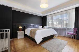 Quarto de hotel moderno com uma cama grande, mesa de cabeceira, uma cadeira e vista para a cidade através de cortinas translúcidas.