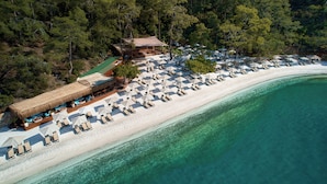 Vista aérea de um resort na praia, com espreguiçadeiras e guarda-sóis, um edifício com telhado de palha e um mar azul-turquesa transparente.