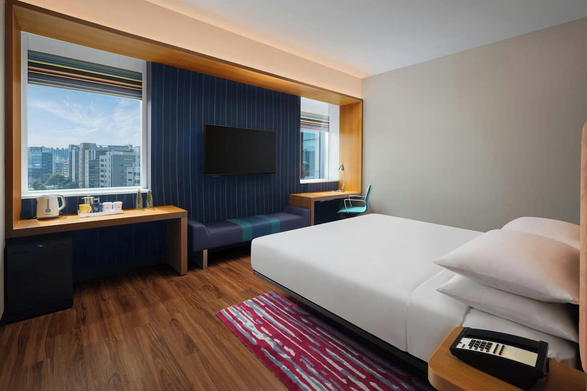 Quarto de hotel moderno com uma cama grande, TV fixada na parede, escrivaninha com cadeira e vista para a cidade através de amplas janelas.