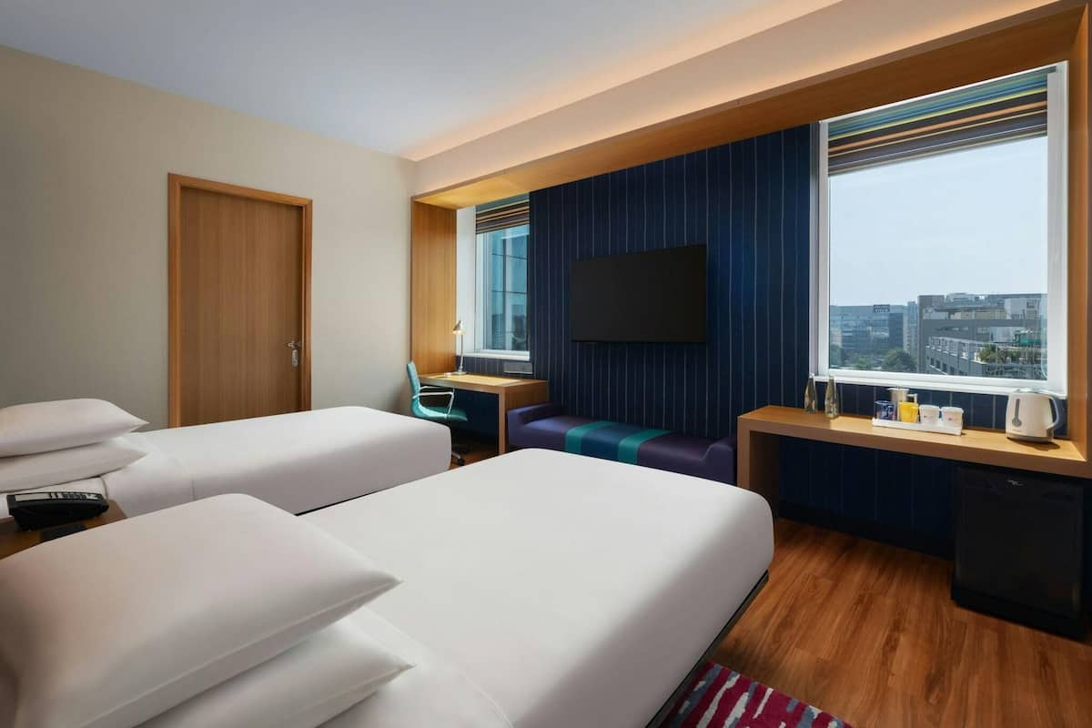Quarto de hotel com duas camas, televisão, escrivaninha com cadeira e janela com vista para a cidade.