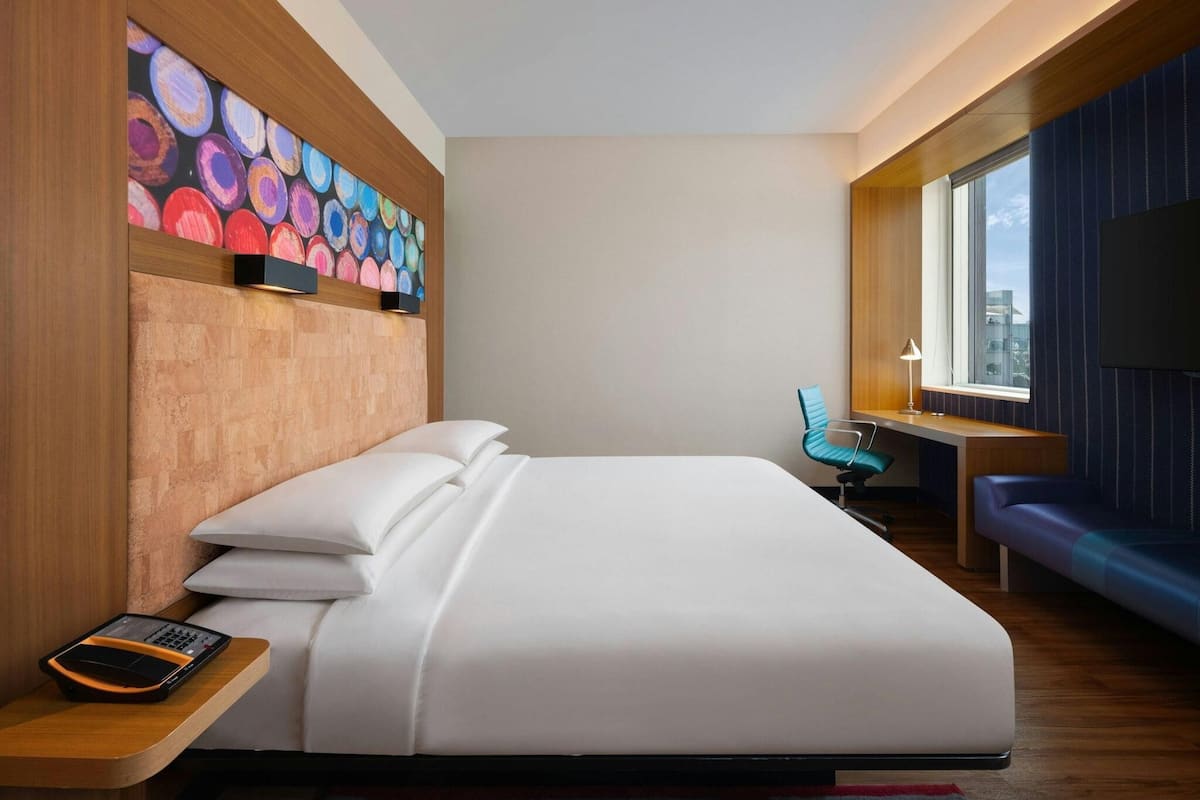 Quarto de hotel moderno com uma cama grande, uma escrivaninha com cadeira, televisão e janela com vista para a cidade.