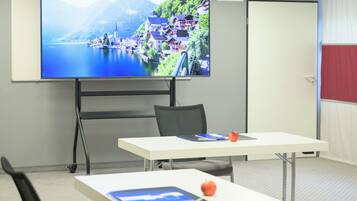 Uma sala de conferências com uma tela grande exibindo uma vista panorâmica, uma mesa branca com um laptop e uma maçã, e uma cadeira preta.