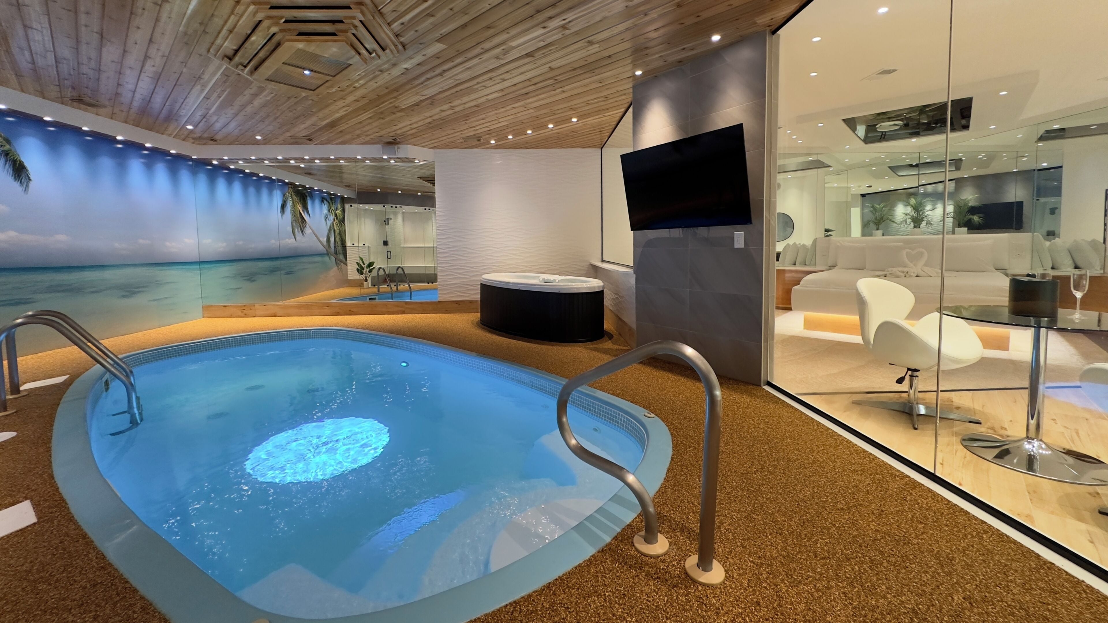 Piscina interna com um mural tropical, hidromassagem e área de estar moderna com parede de vidro.