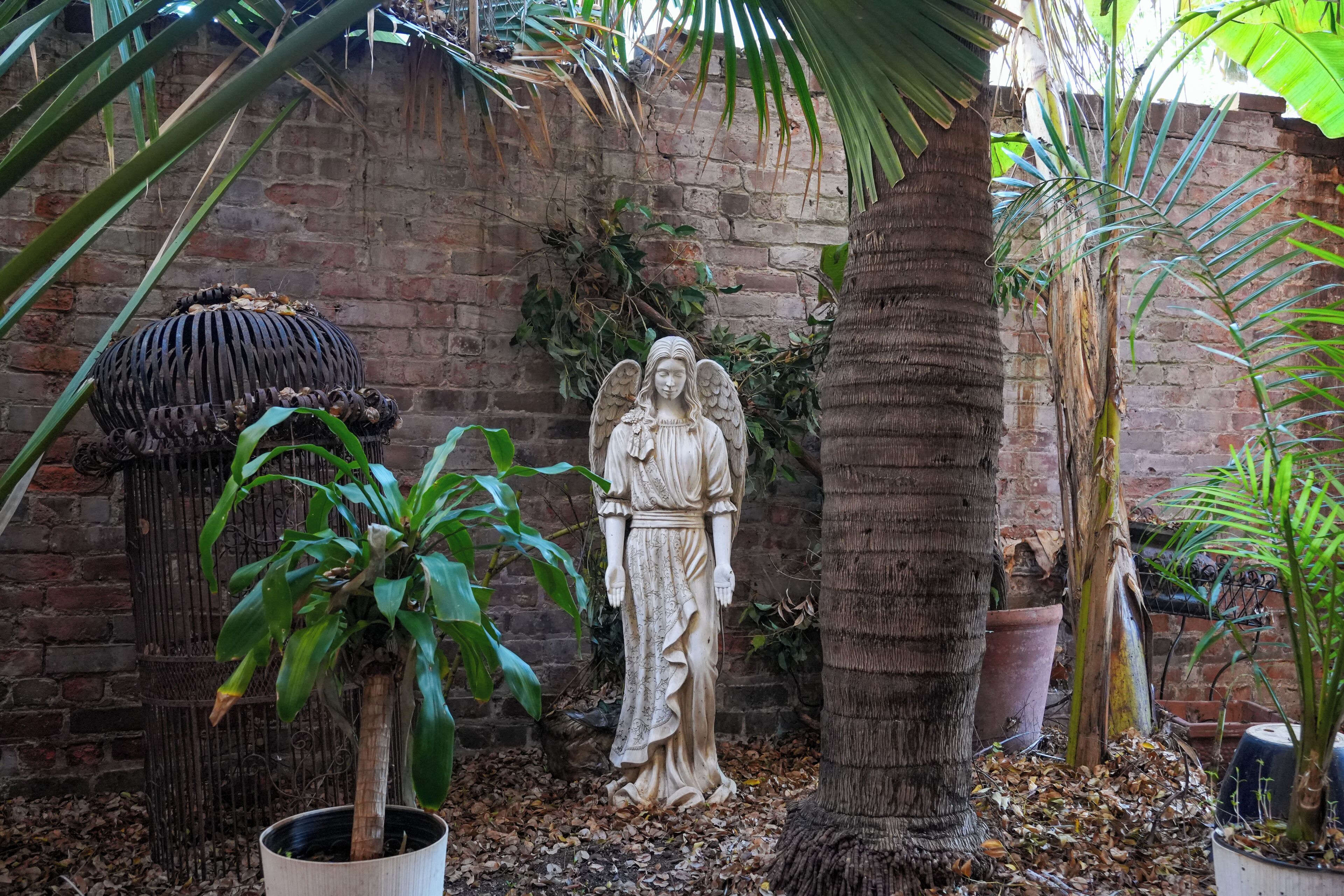 Estátua de um anjo em um jardim com parede de tijolos, vasos de plantas e uma palmeira.