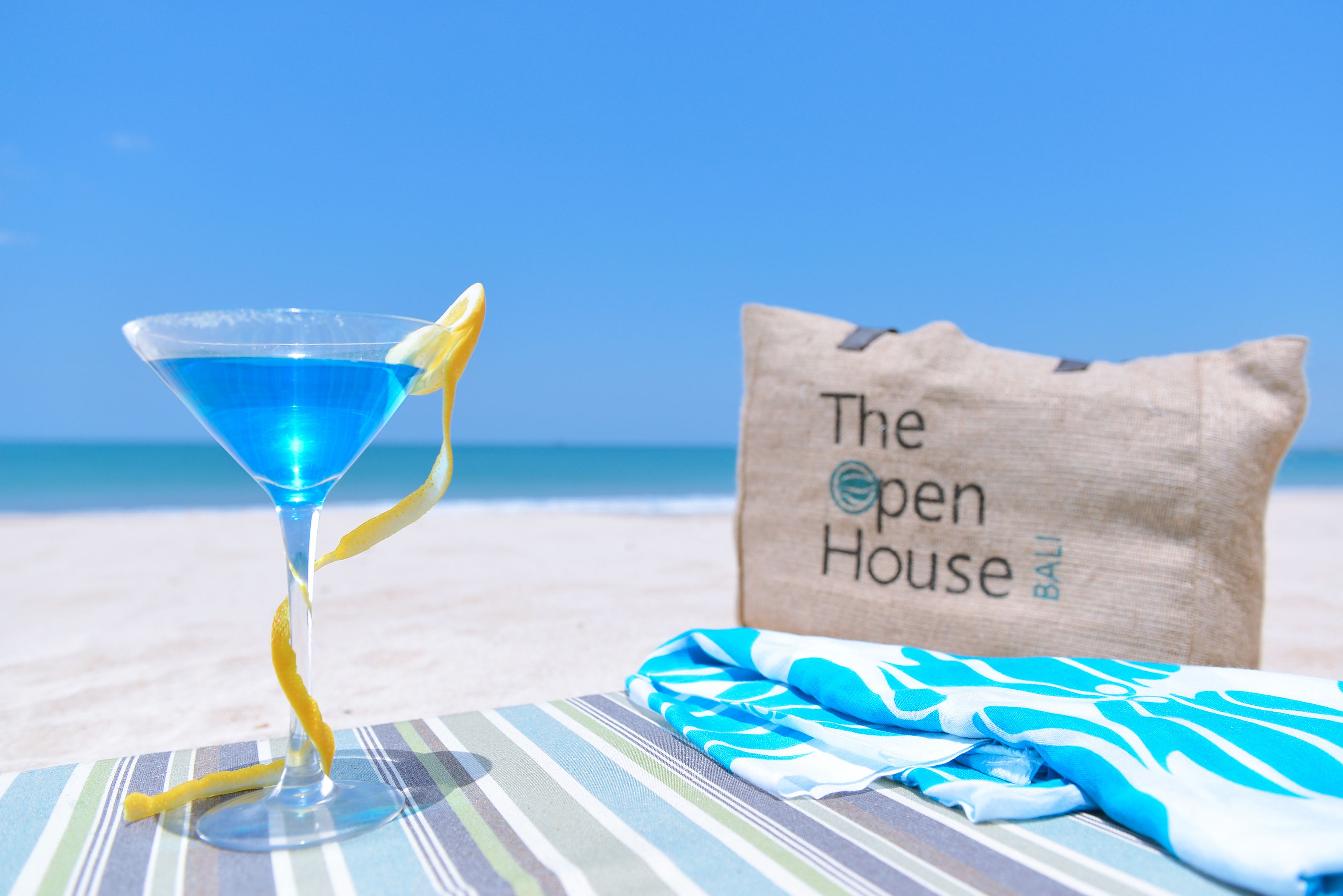 Um copo de martini com uma casca de limão sobre uma toalha de praia listrada, uma bolsa de praia com a etiqueta "The Open House Bali" e um céu azul límpido.
