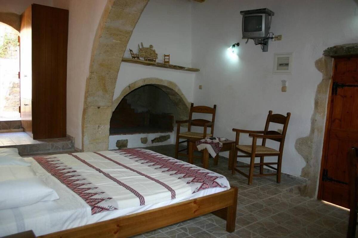 Quarto com uma cama, duas cadeiras de madeira, lareira de pedra e uma televisão fixada na parede.