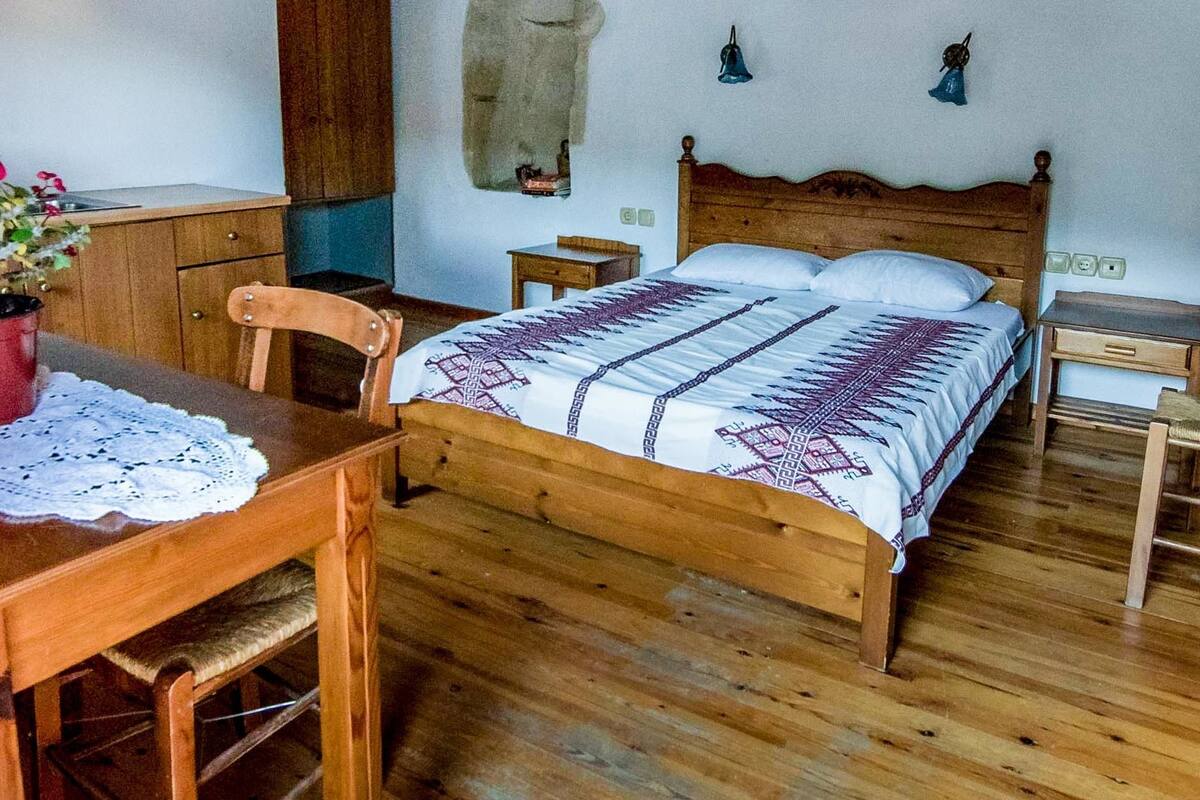 Quarto com piso de madeira, uma cama, uma mesinha de cabeceira de madeira, uma pequena mesa com toalha de renda e uma cadeira de madeira.