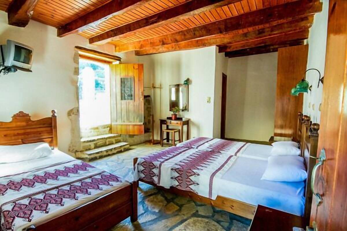 Quarto com duas camas de madeira, soalho de pedra, teto de madeira e uma pequena mesa com uma cadeira.
